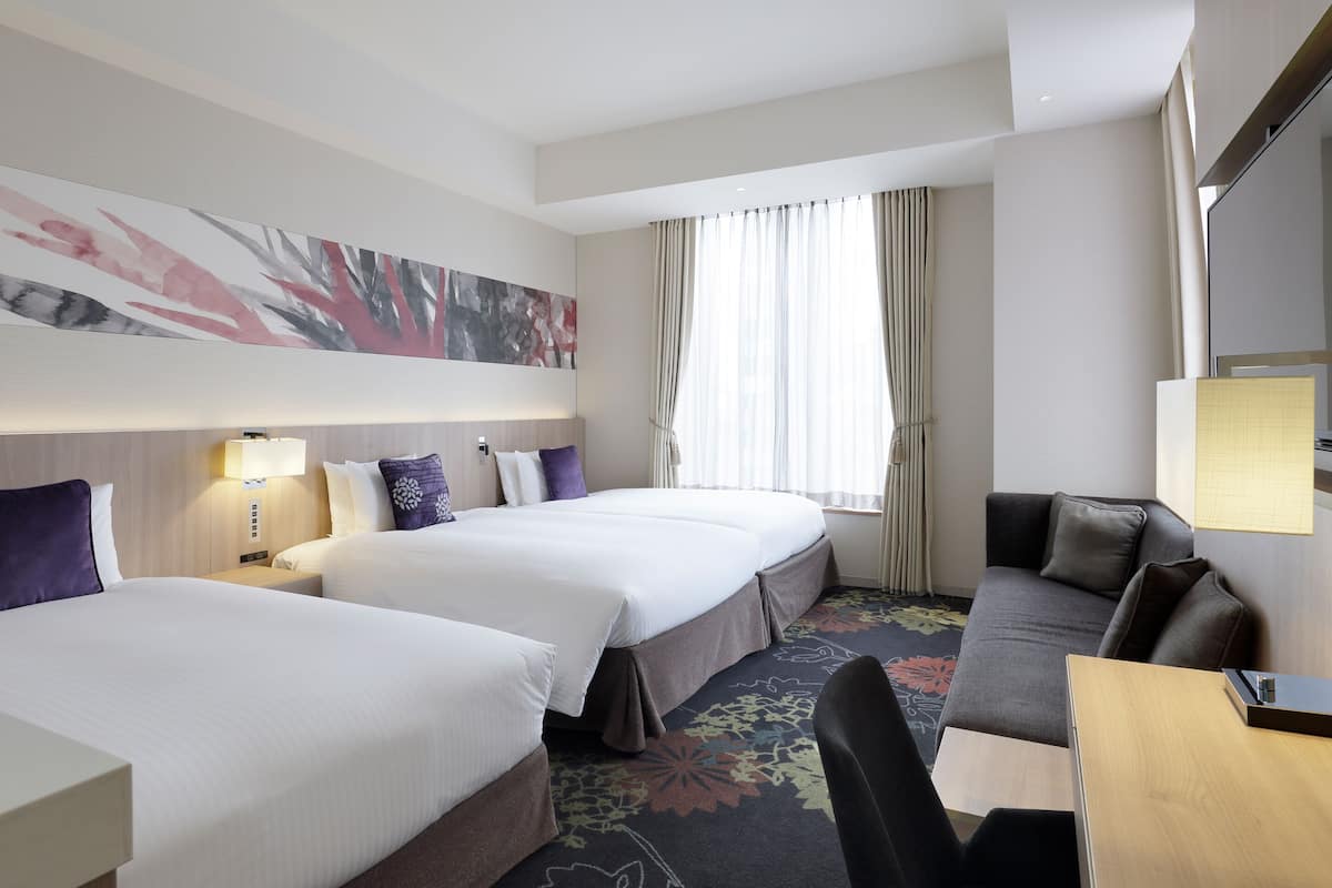 Quarto de hotel com duas camas, um sofá, uma escrivaninha e uma janela grande com cortinas.