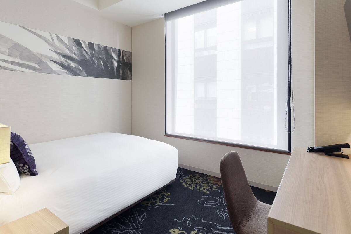 Quarto de hotel com cama, escrivaninha, cadeira, janela e obra de arte na parede.