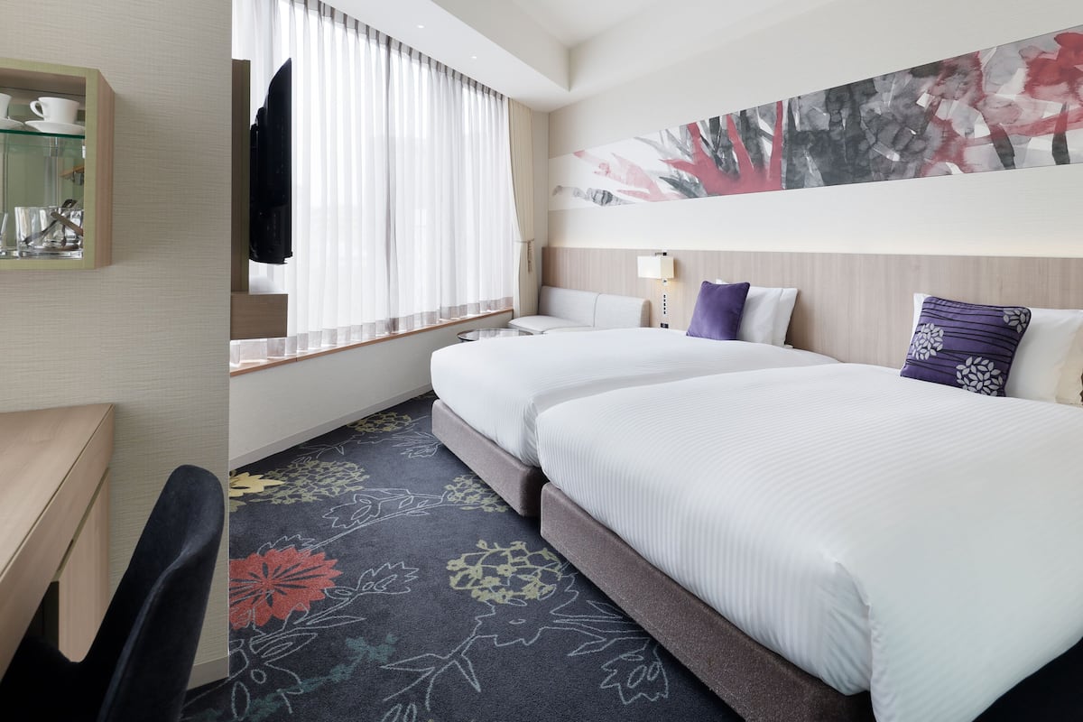 Quarto de hotel com duas camas, uma escrivaninha, uma cadeira, uma televisão e um quadro abstrato grande na parede.