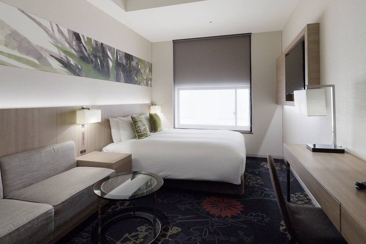 Quarto de hotel com uma cama, um sofá, uma mesinha, uma televisão e um quadro na parede.