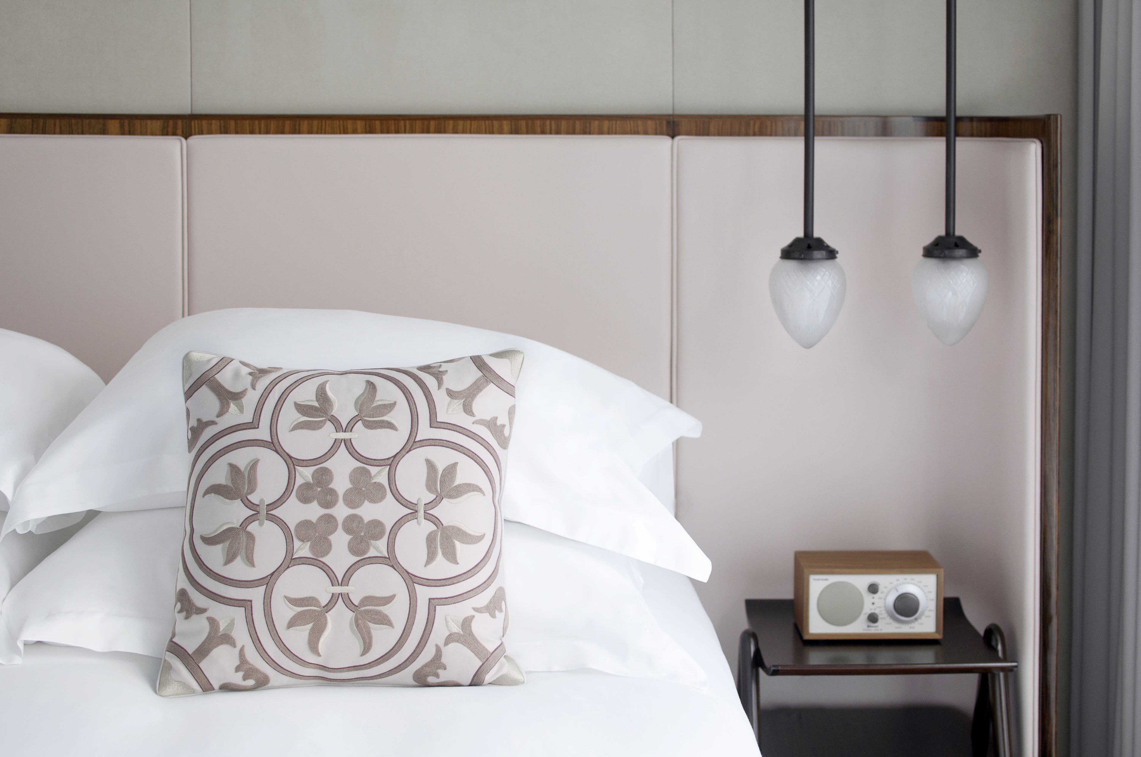 Cama bem arrumada com lençóis brancos e almofada estampada, mesa de cabeceira com rádio e cabeceira com painel de madeira.