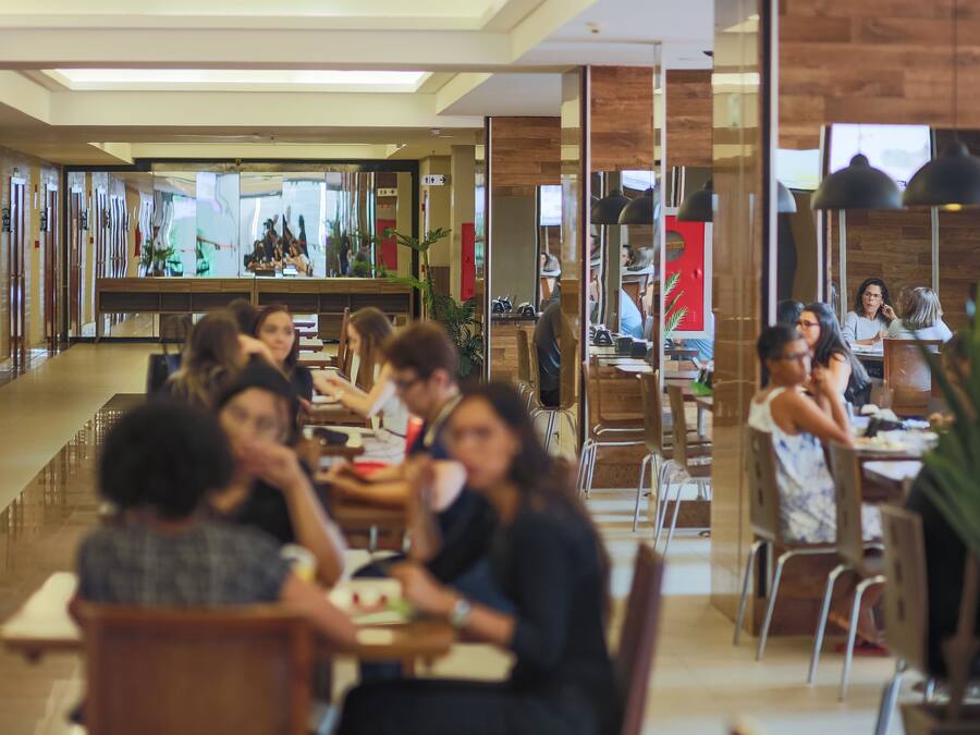 Uma sala de jantar moderna com várias mesas e cadeiras, um balcão comprido e diversos vasos com plantas.