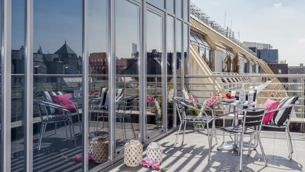Terraço no telhado com paredes de vidro, cadeiras de metal e uma mesa posta com flores e uma cesta de frutas.