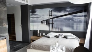 Quarto de hotel moderno com uma cama grande, uma mesa de cabeceira, uma mesa de vidro com duas taças de vinho e um mural grande na parede com a imagem de uma ponte.