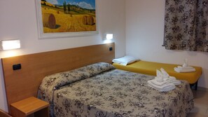 Quarto de hotel com cabeceira de madeira, duas camas com roupa de cama florida, um quadro de paisagem rural e uma janela com cortinas florais.