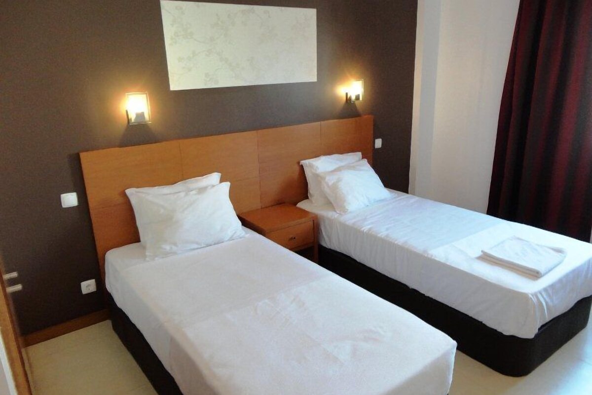 Quarto de hotel com duas camas de solteiro, cabeceira de madeira, mesa de cabeceira e luminária fixada na parede.