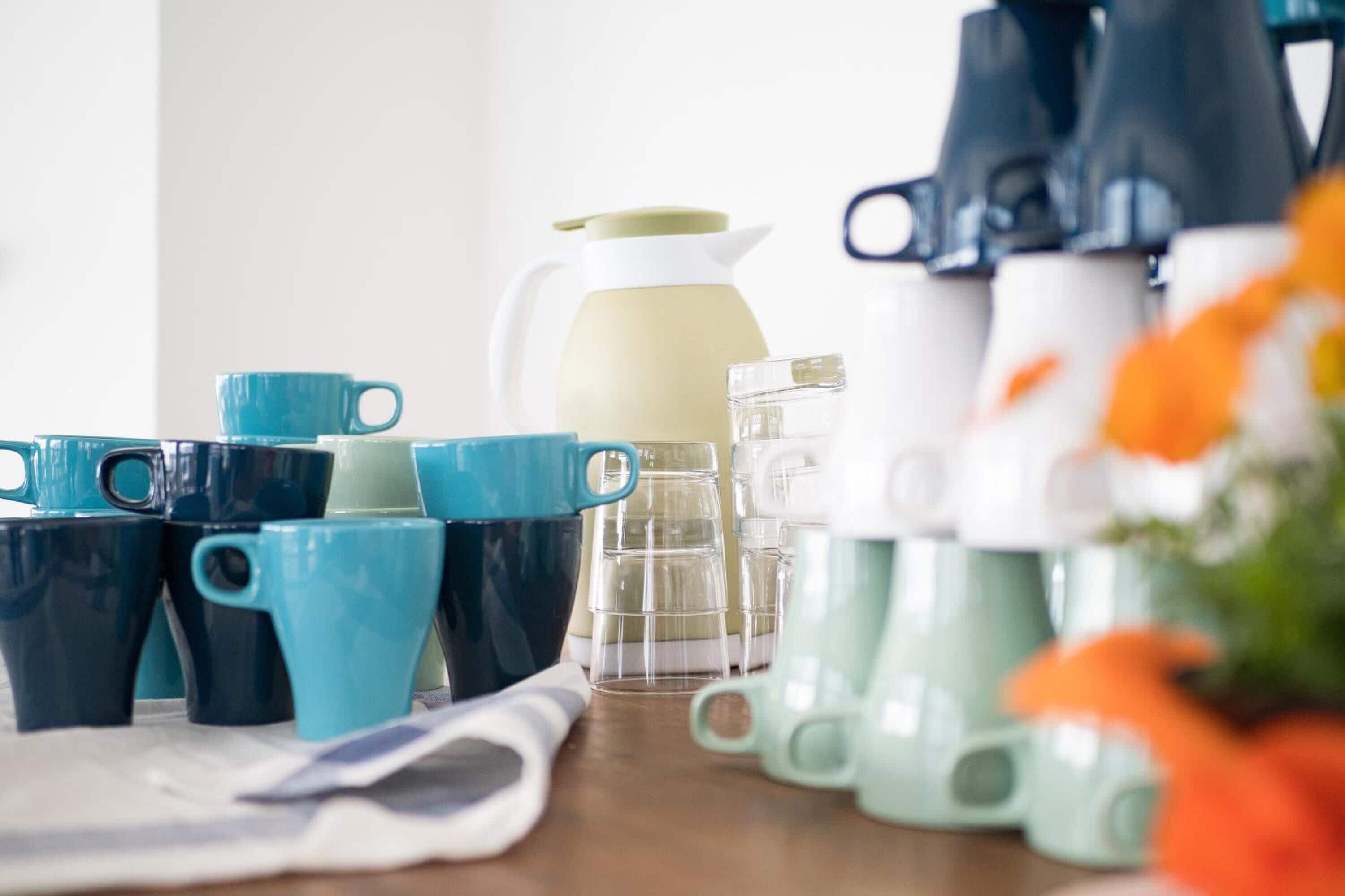 Conjunto de utensílios de cozinha incluindo chávenas azuis e verdes, um jarro branco e copos de vidro transparente sobre uma mesa de madeira.