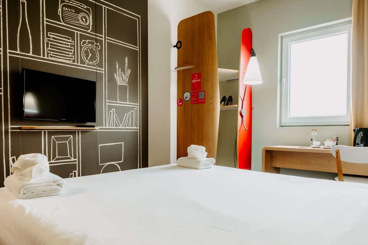 Quarto de hotel com cama, televisão, guarda-roupa de madeira, uma prancha de surf vermelha, escrivaninha e janela.