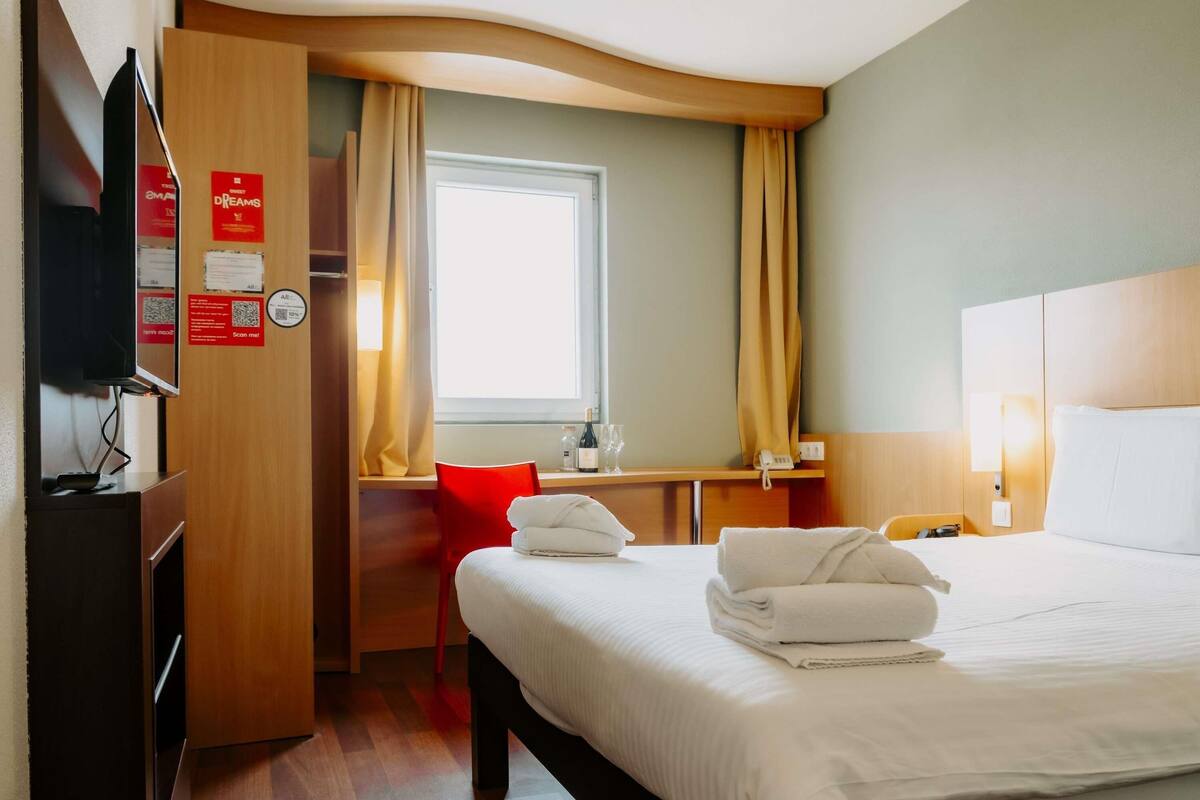 Quarto de hotel com cama, escrivaninha, cadeira, televisão e janela com cortinas.