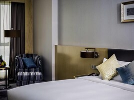 Quarto de hotel com cama, uma cadeira, luminária na parede e uma mesa com frutas.