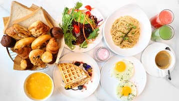 Uma mesa de café da manhã com waffles, ovos, torradas, doces, salada, macarrão e diversas bebidas.