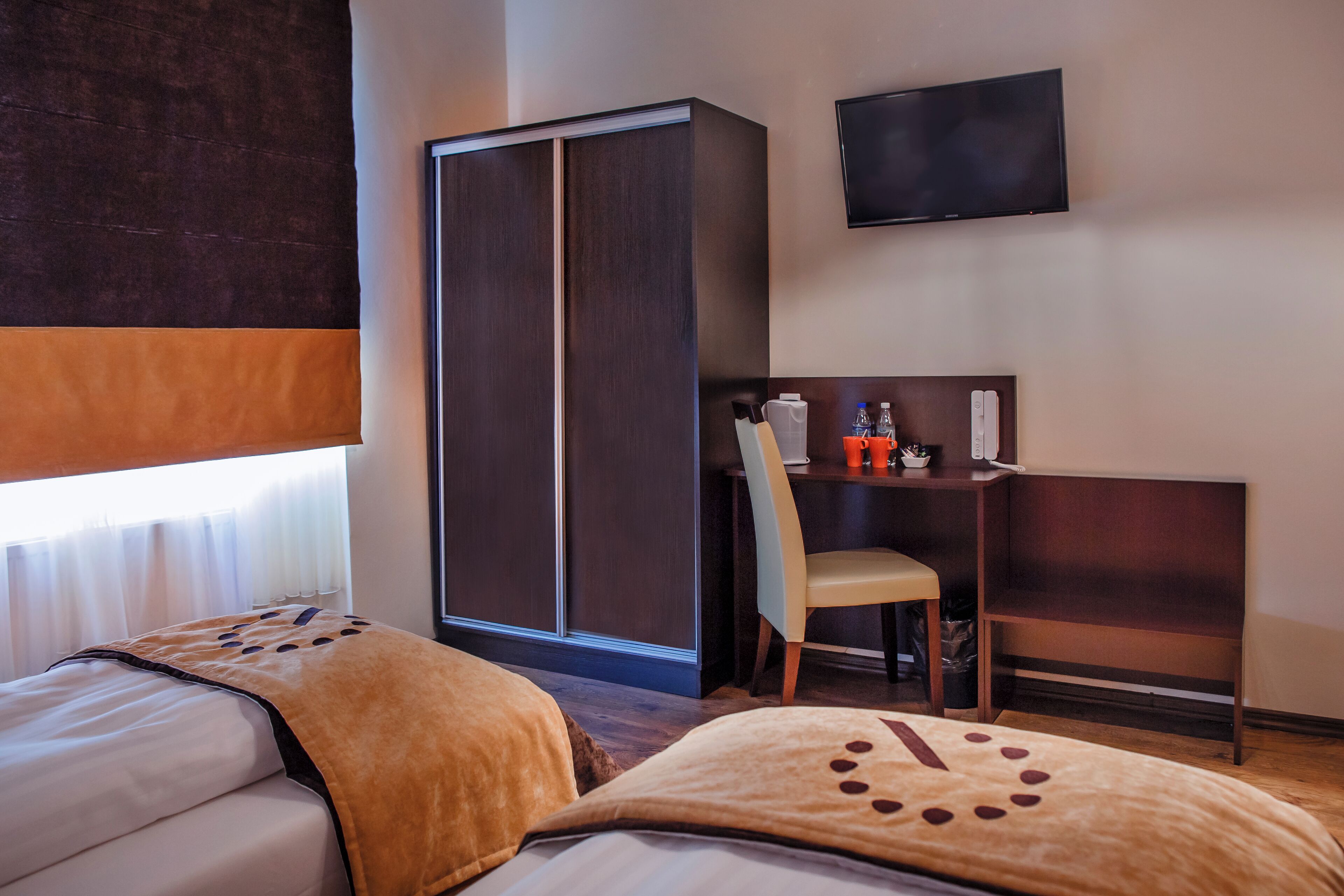 Quarto de hotel com duas camas, um guarda-roupa, uma secretária com cadeira, televisão e janela com cortinas.