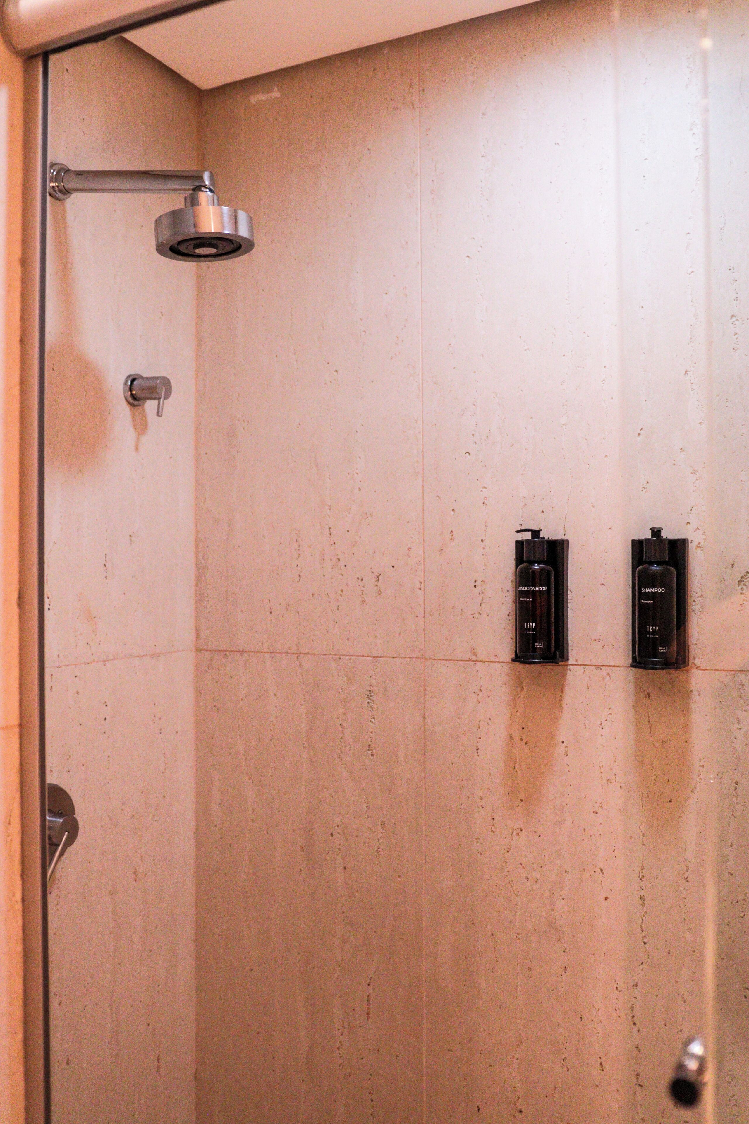 Área de chuveiro com chuveiro manual, painel de controle e duas embalagens de shampoo fixadas na parede.