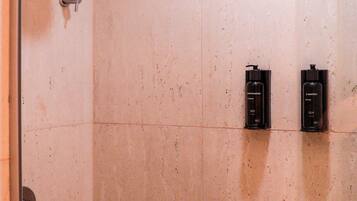 Área de chuveiro com chuveiro manual, painel de controle e duas embalagens de shampoo fixadas na parede.