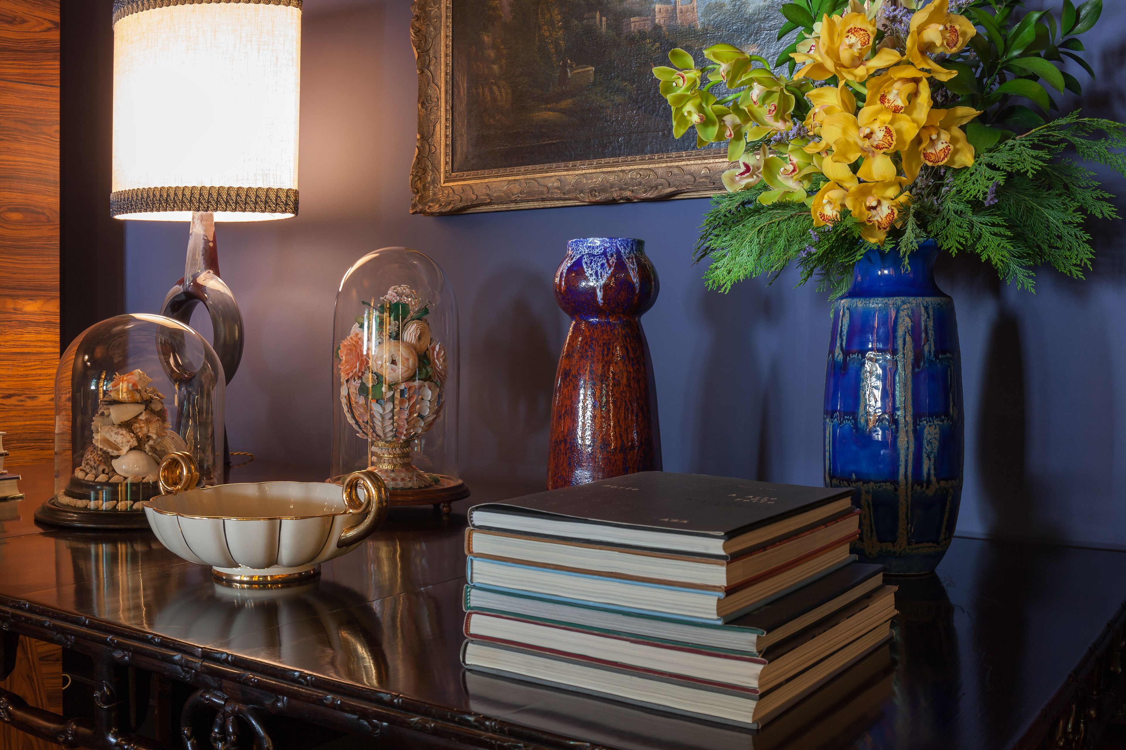 Uma mesa com uma luminária, um vaso com flores, uma tigela decorativa e uma pilha de livros.