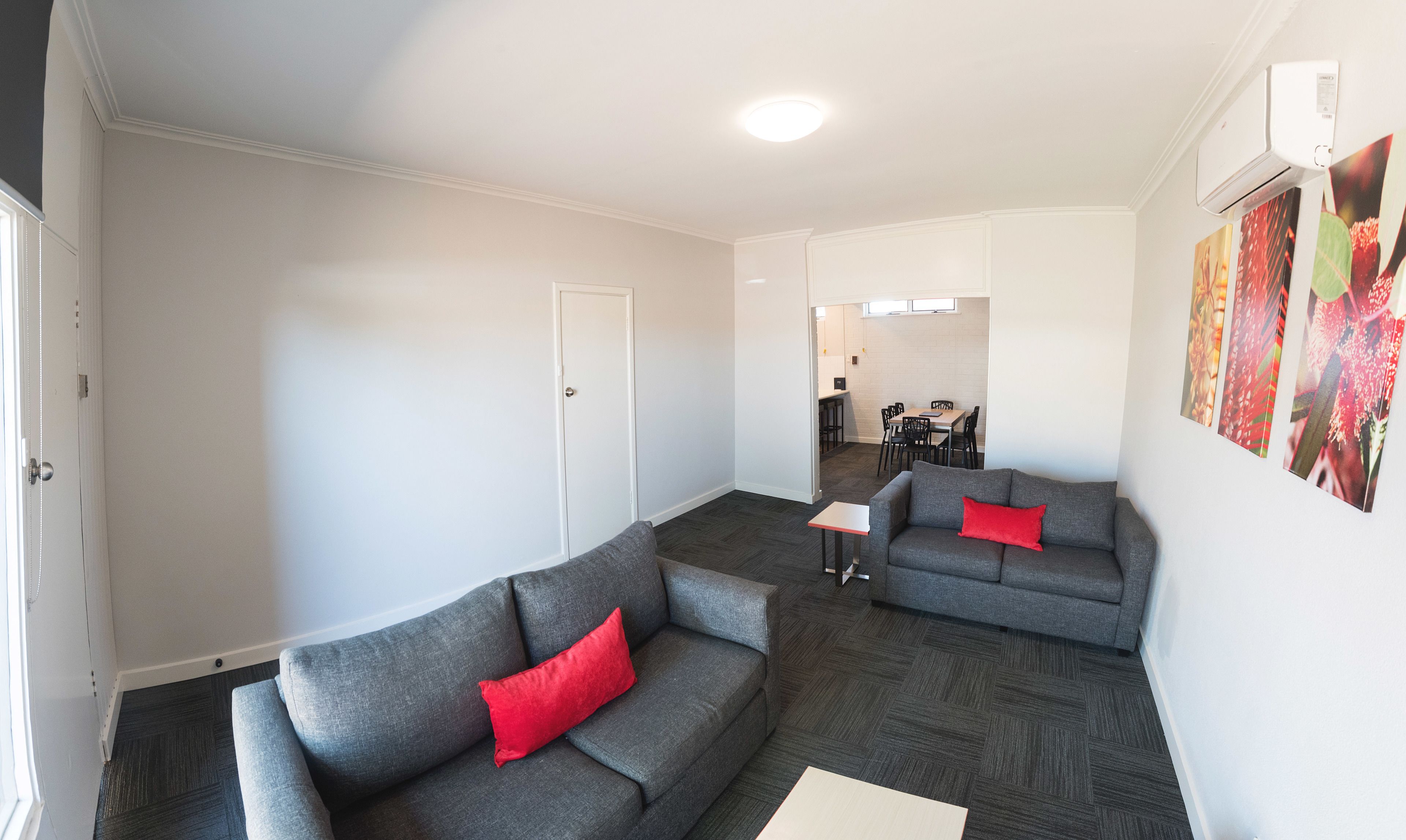 Uma sala de estar moderna com dois sofás cinzas, cada um com uma almofada vermelha, e uma pequena mesa branca. O espaço inclui uma porta branca, uma parede branca e uma área de jantar ao fundo.