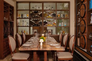 Uma sala de jantar com uma mesa comprida posta para seis pessoas, rodeada por cadeiras de madeira. Atrás da mesa, há prateleiras com garrafas de vinho e copos.