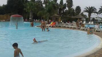 Piscina com pessoas aproveitando a água, cercada por palmeiras e cadeiras de descanso.