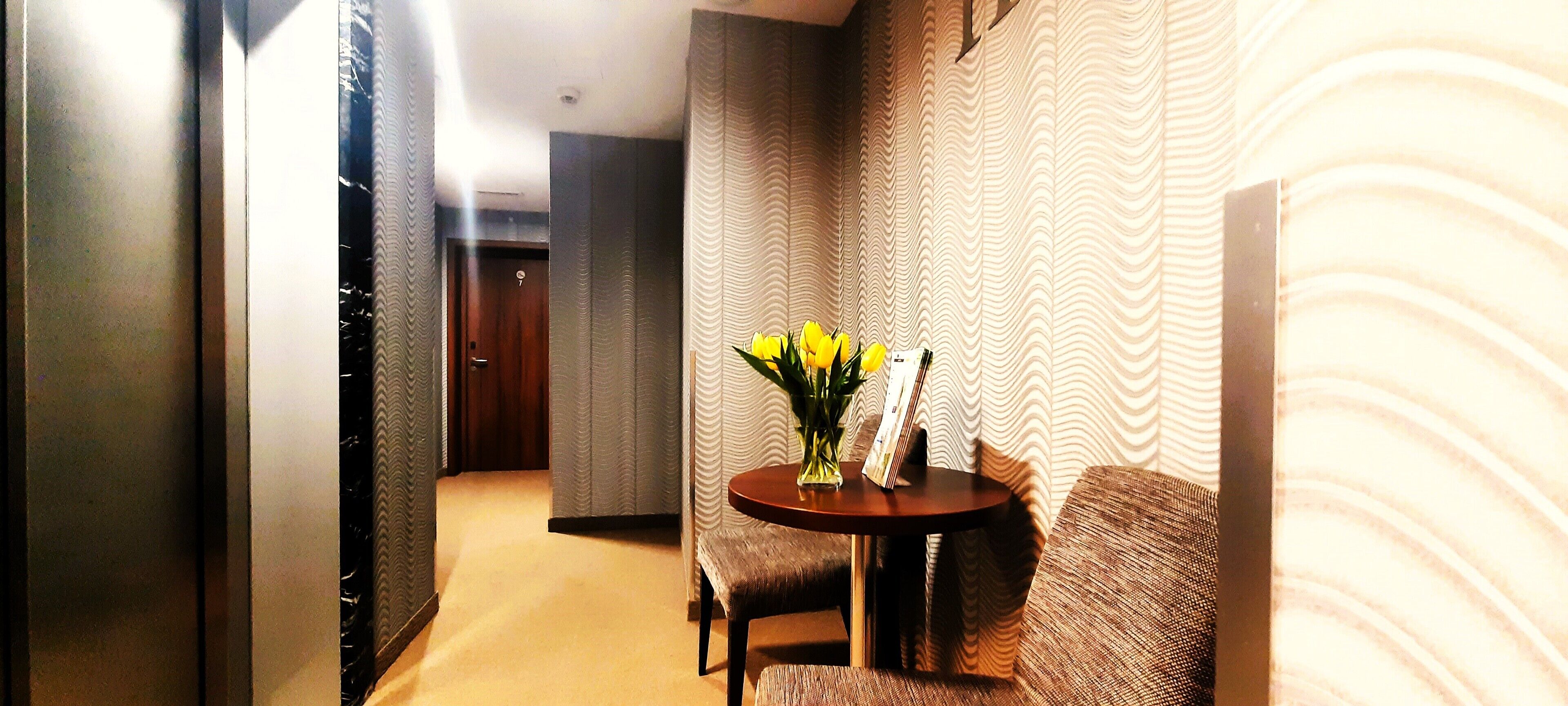 Um corredor de hotel com uma mesa redonda, um vaso com flores amarelas e um sofá.
