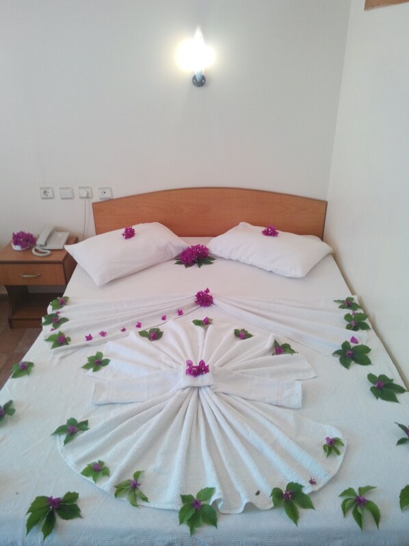 Uma cama bem arrumada com arranjo decorativo de toalhas e flores roxas, um telefone ao lado da cama e um luminária fixada na parede.