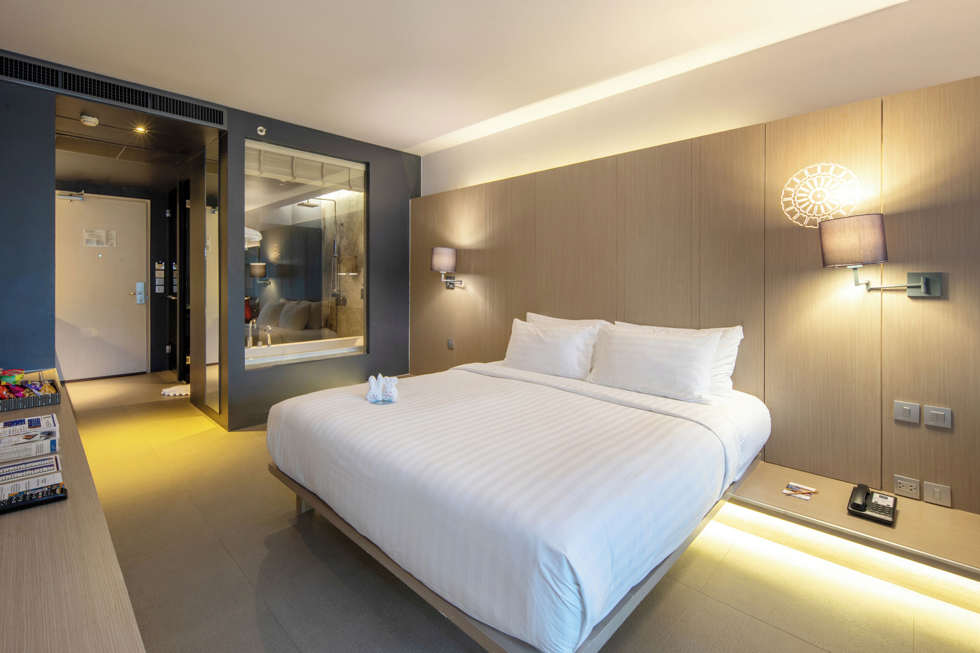Quarto de hotel moderno com uma cama grande, abajures, um espelho e mesa de cabeceira com telefone e revistas.