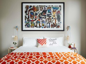Uma cama bem arrumada com um edredom estampado, uma pintura abstrata emoldurada acima dela e mesas de cabeceira com luminárias e pequenas plantas.