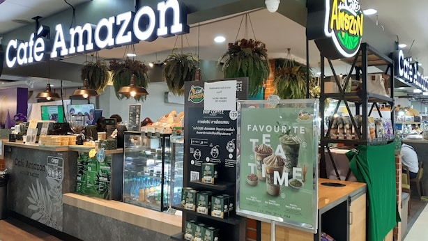 Um café chamado 'Café Amazon' com uma placa, balcão exibindo bebidas e plantas penduradas.