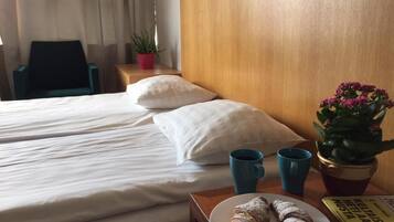 Quarto de hotel com duas camas, cabeceira de madeira, uma mesinha com croissants, uma planta em vaso e uma revista.