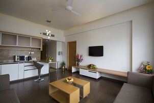 Uma sala de estar moderna com um sofá, uma mesa de centro com frutas, uma televisão e uma cozinha com micro-ondas.