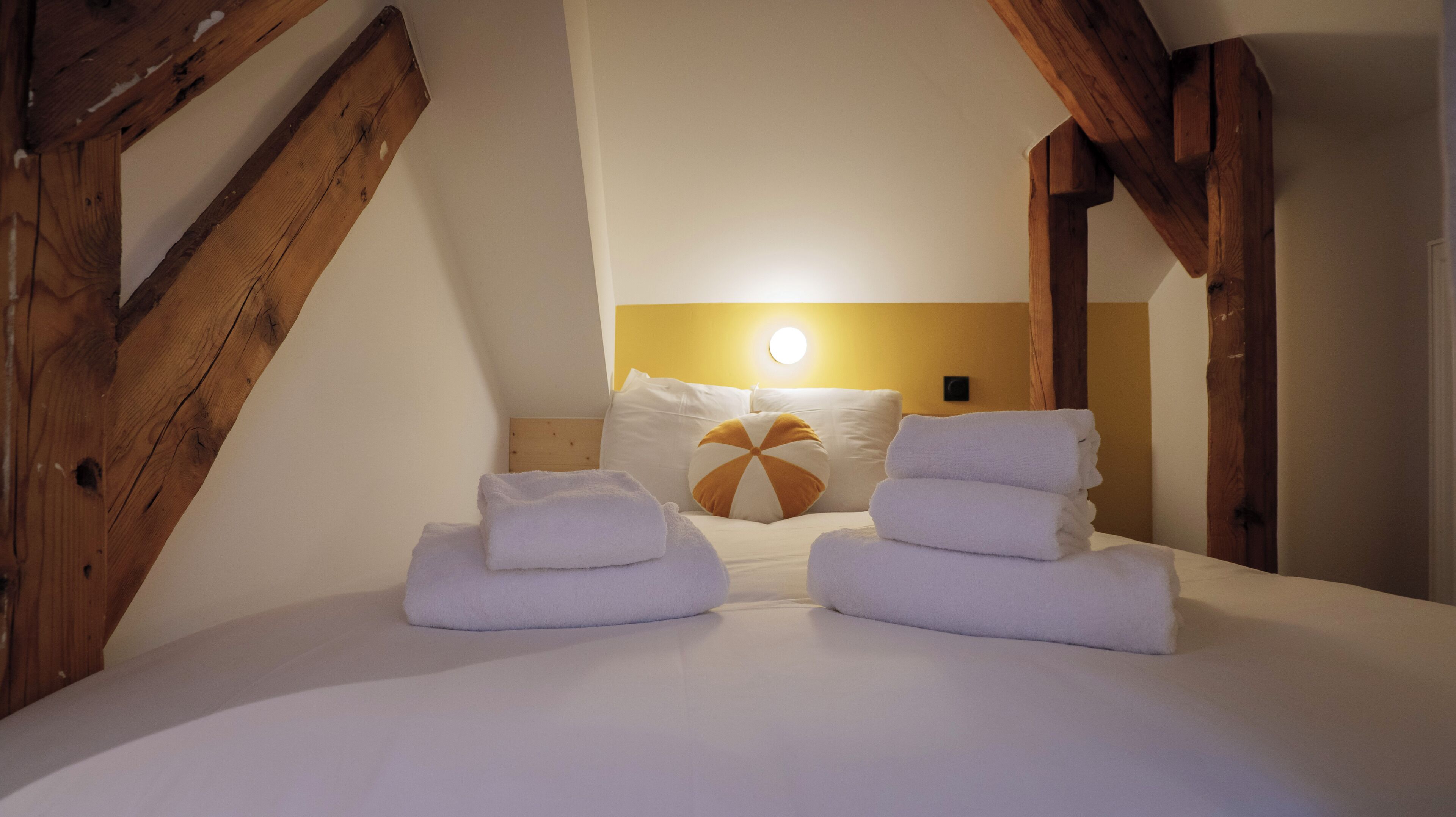 Quarto de hotel com teto de madeira, cama com lençóis brancos, parede amarela de destaque e travesseiro redondo laranja e branco.