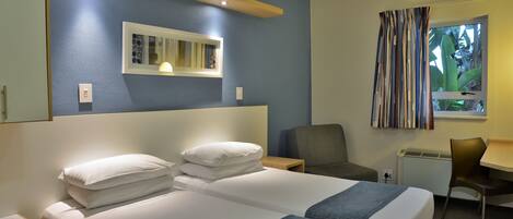 Quarto de hotel com duas camas, uma escrivaninha, uma cadeira, uma janela com cortinas e uma prateleira na parede com espelho e iluminação.