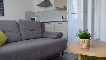 Uma sala de estar moderna com um sofá cinzento, uma mesa redonda pequena com uma planta, e uma cozinha ao fundo.