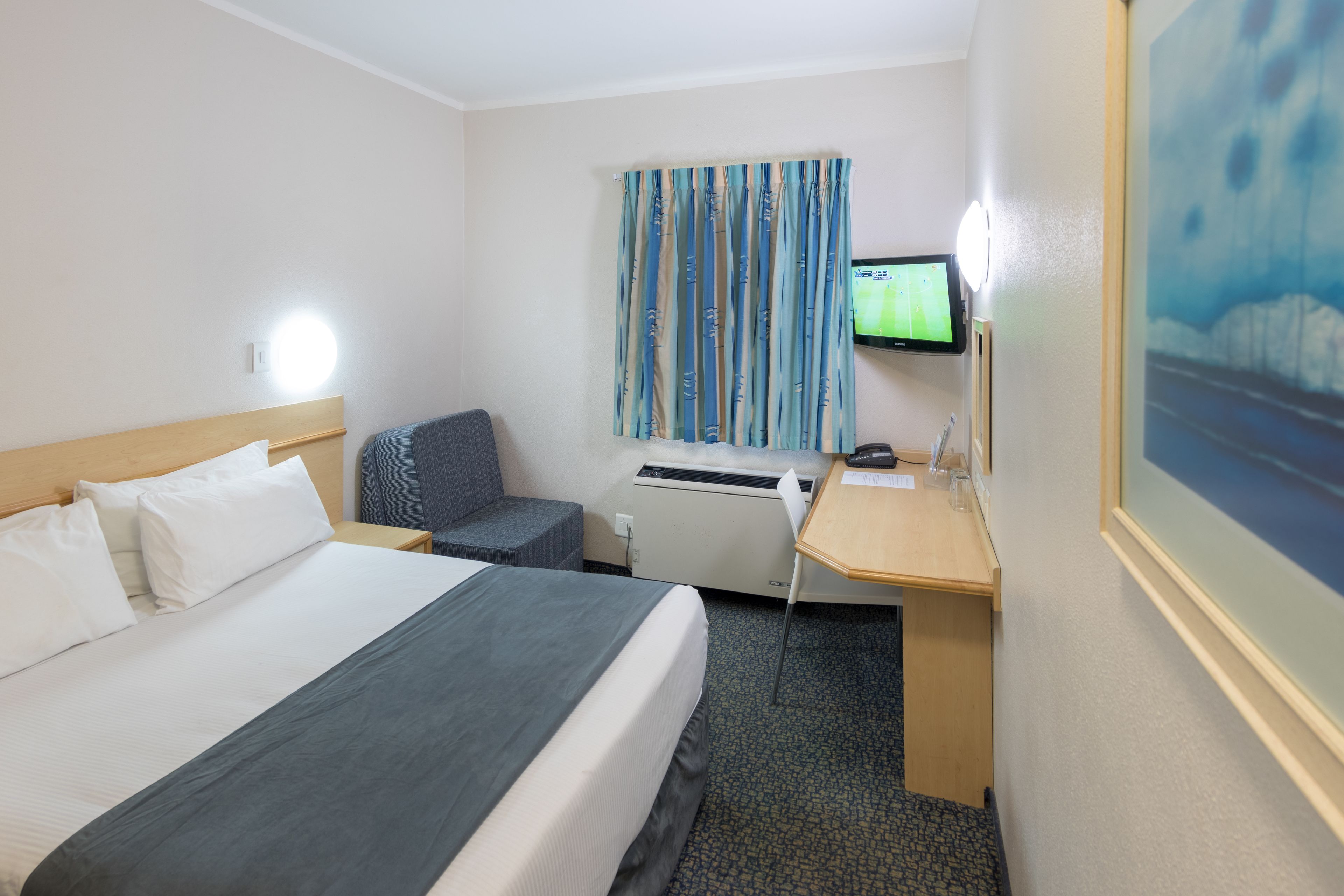 Quarto de hotel com cama, escrivaninha, cadeira, televisão e janela com cortinas.