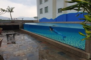 Uma piscina com uma pessoa nadando, um deck com ladrilhos de pedra e um edifício de paredes azuis e brancas.