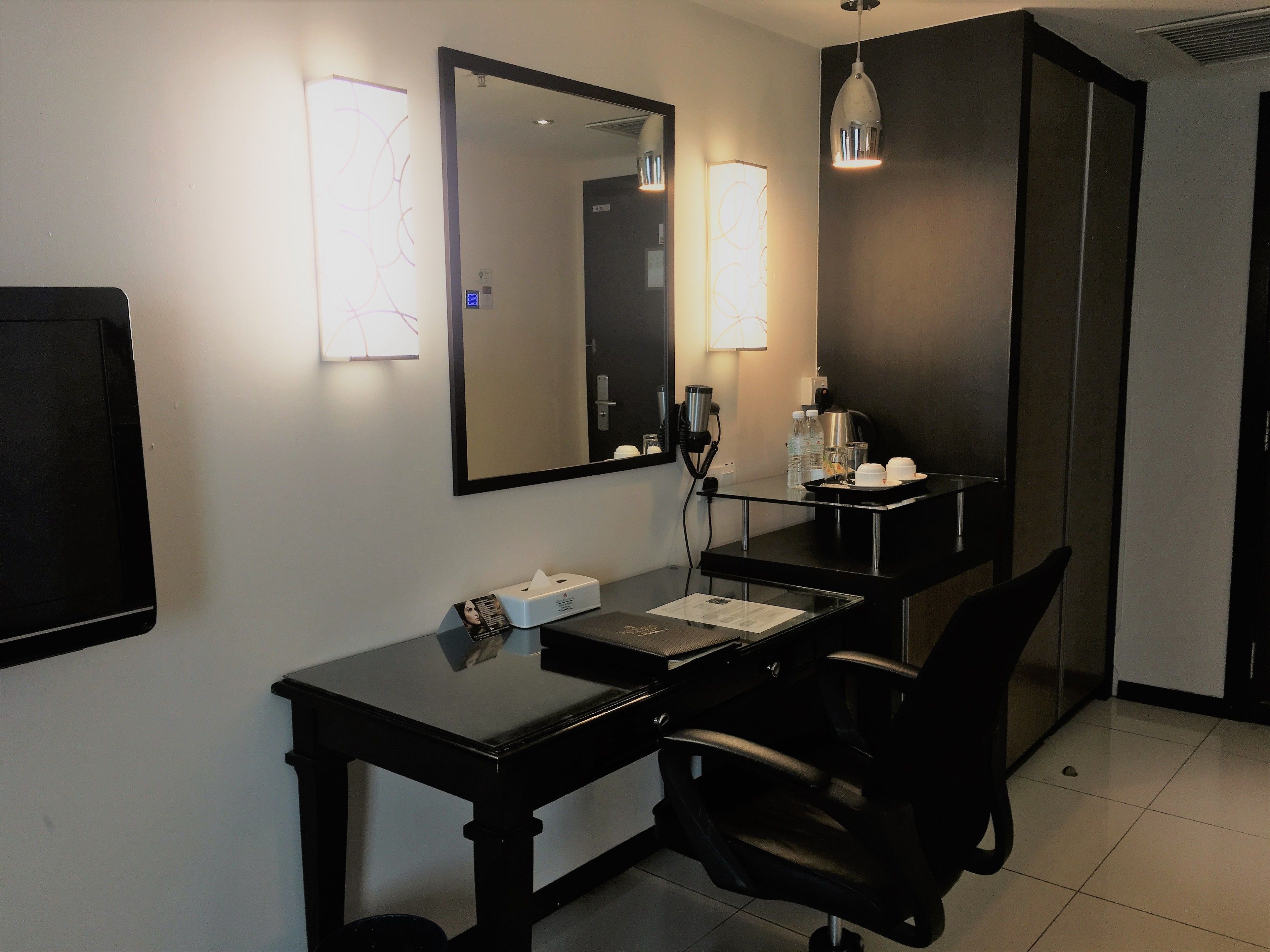 Quarto de hotel com escrivaninha, cadeira e um espelho grande. Há uma luminária na escrivaninha e uma garrafa de água no balcão.
