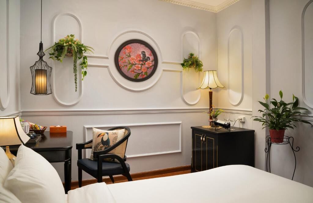 Quarto de hotel com cama, escrivaninha com luminária, cadeira, armário e plantas suspensas na parede.