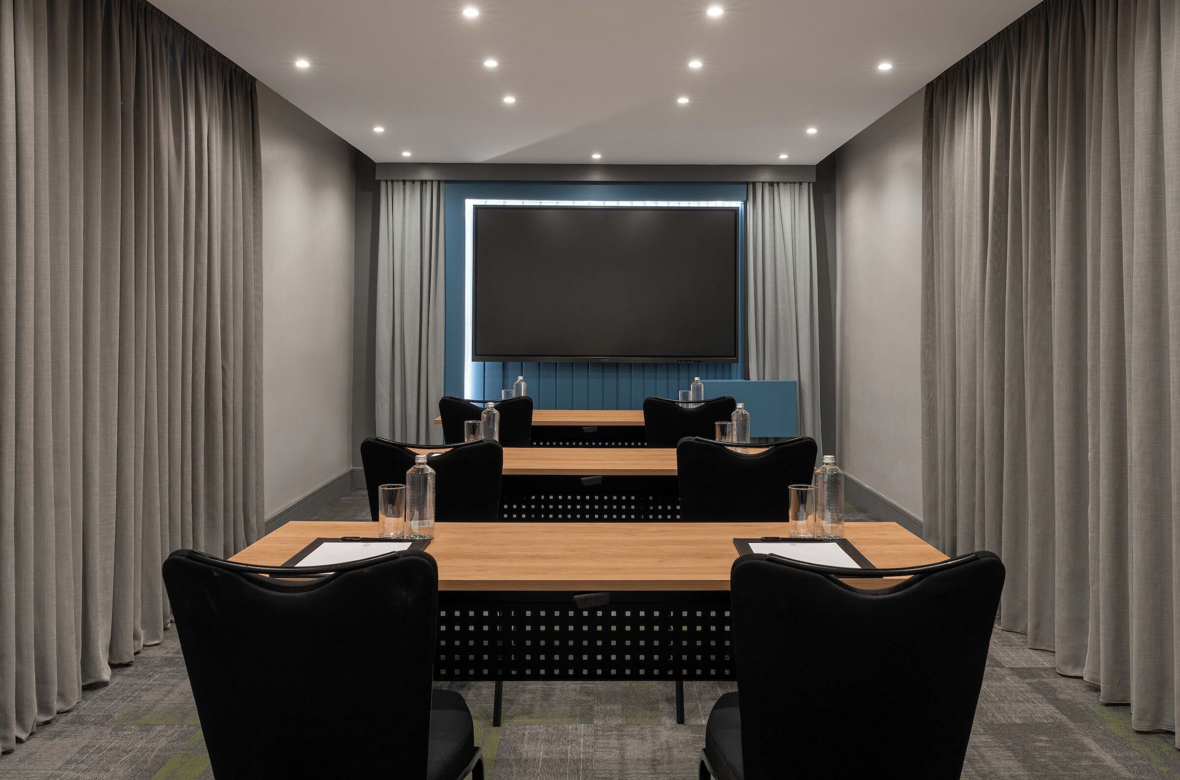 Uma sala de conferências com uma mesa grande, cadeiras pretas e uma tela grande na frente.