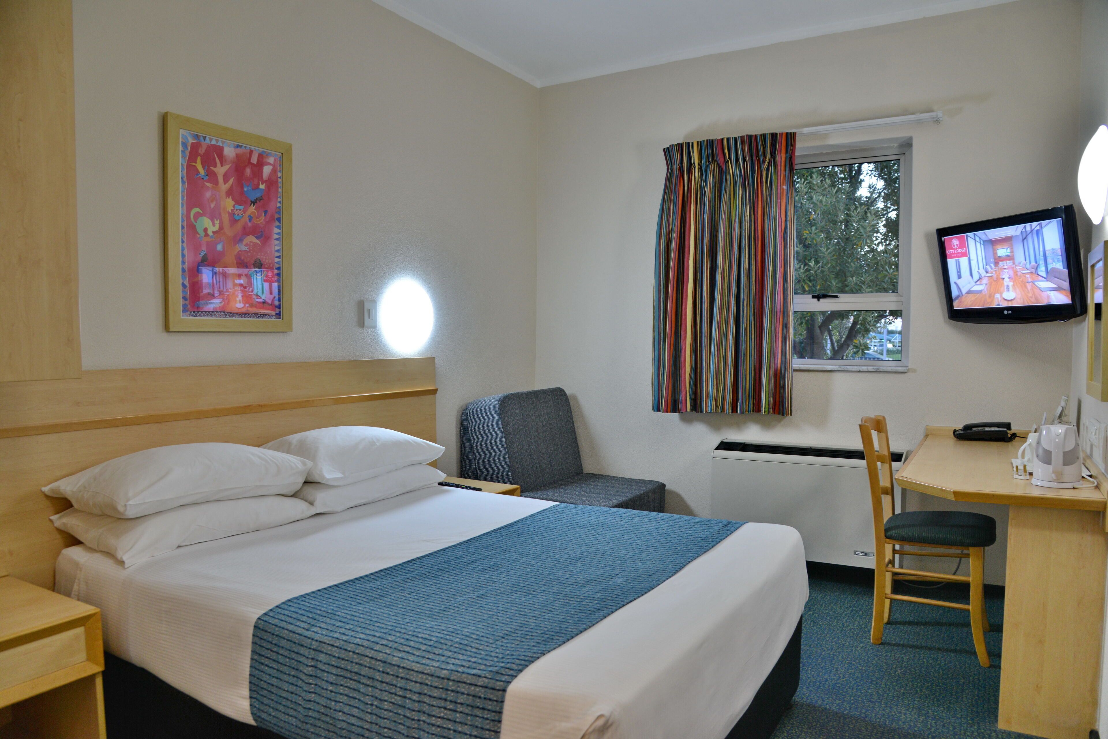 Quarto de hotel com cama, escrivaninha, cadeira, televisão e janela com cortinas.
