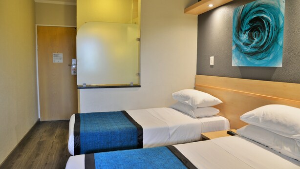 Quarto de hotel com duas camas, uma cabeceira de madeira, um quadro abstrato grande e um espaço com chuveiro.