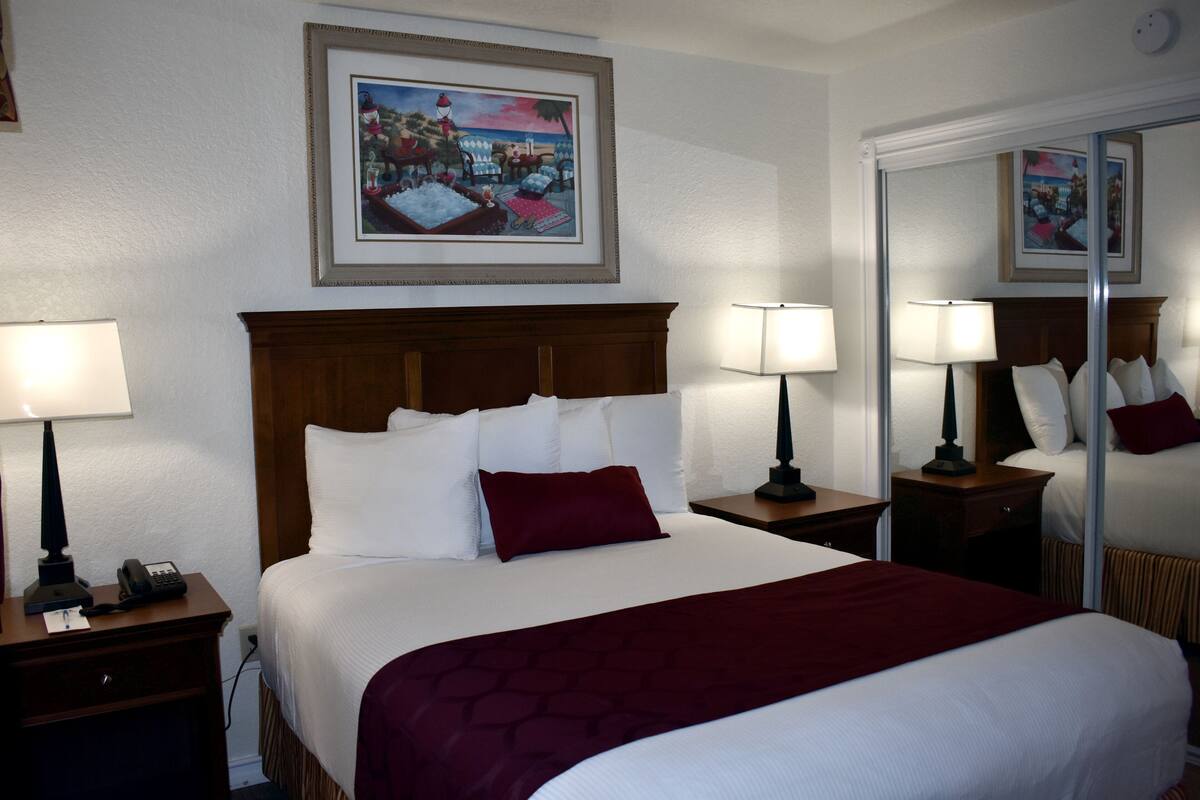 Quarto de hotel com uma cama grande, duas mesas de cabeceira com luminárias, um criado-mudo com telefone e um quadro emoldurado acima da cama.