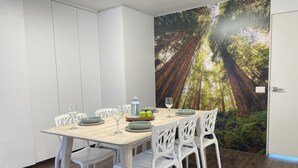 Uma área de refeições com uma mesa de madeira posta para seis pessoas, cadeiras brancas e um mural na parede com tema florestal.