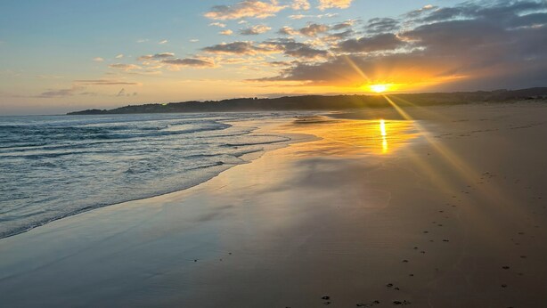 Uma praia com pôr do sol, ondas calmas e céu claro.