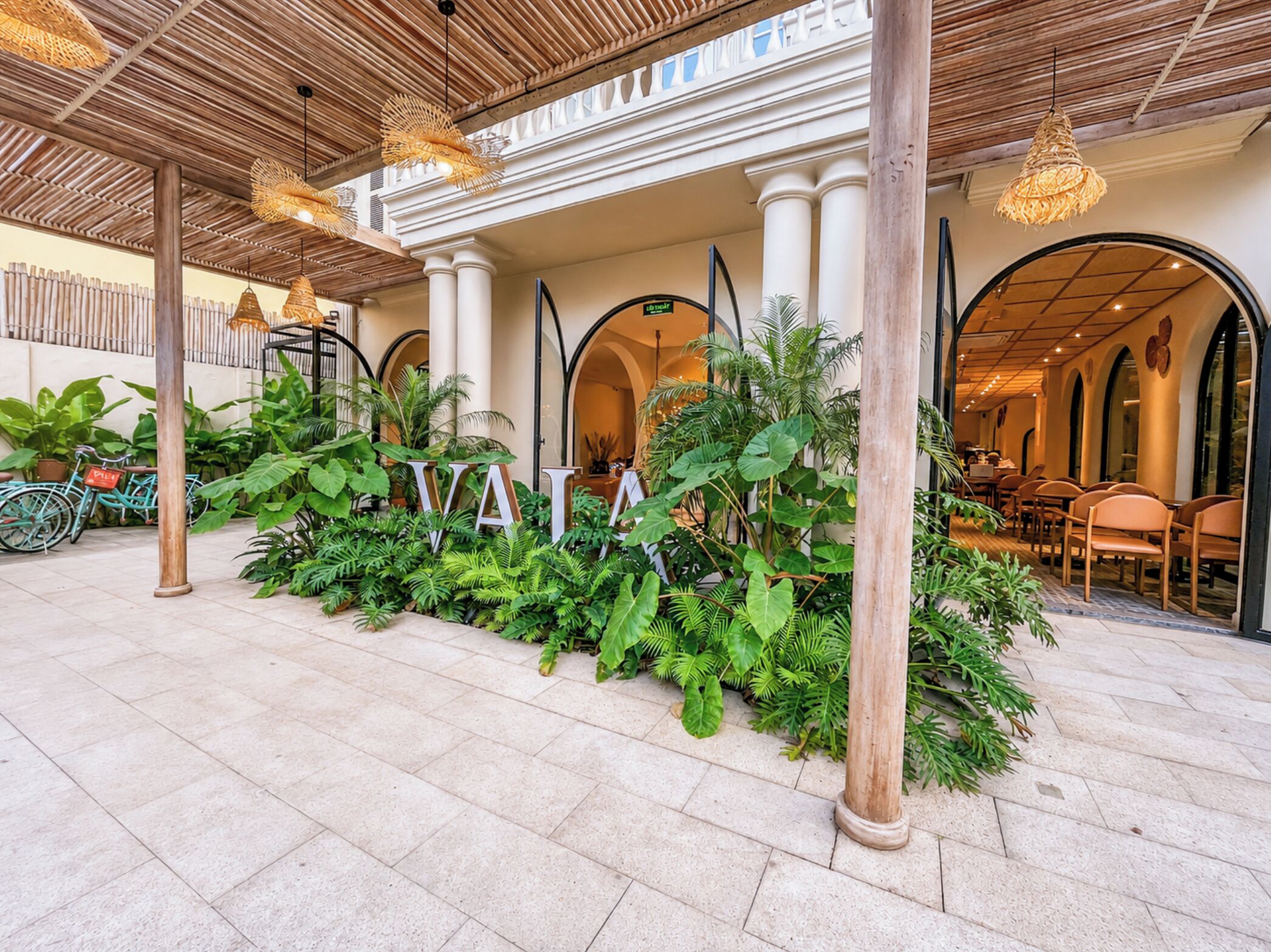 Ampla área externa com piso de azulejos, cercada por plantas verdes exuberantes e colunas de madeira.
