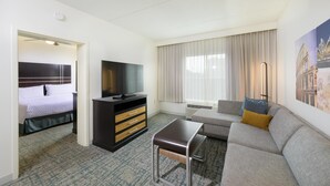 Quarto de hotel com cama, televisão, sofá, mesinha e uma grande janela com cortinas.