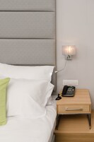 Uma cama bem arrumada com travesseiros brancos e verdes, uma mesinha de cabeceira com luminária, telefone e uma gavetinha.