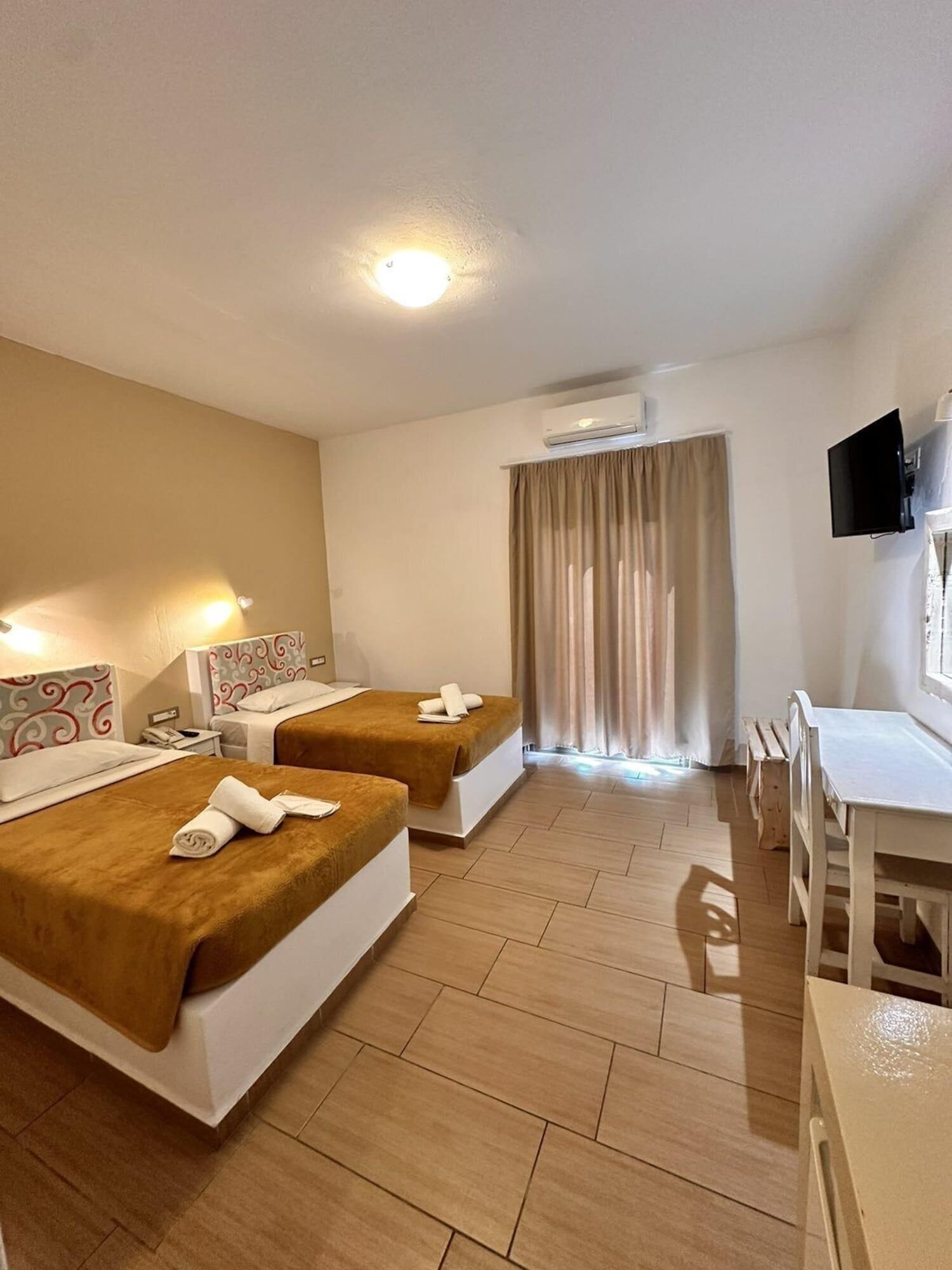 Quarto de hotel com duas camas, uma escrivaninha, uma cadeira, ar condicionado, televisão e janela com cortinas.