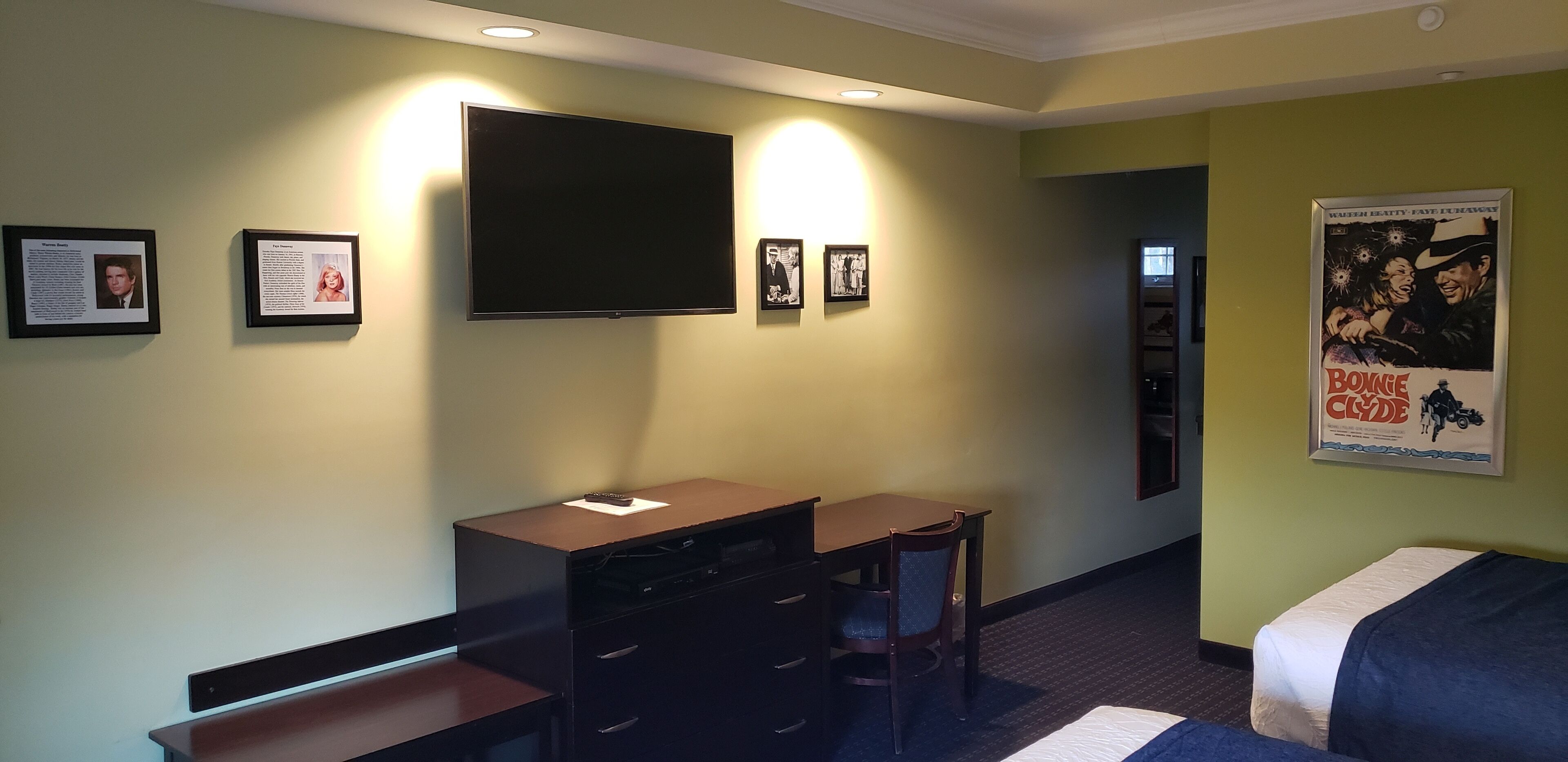 Quarto de hotel com televisão de tela plana na parede, escrivaninha com cadeira, cama e quadros nas paredes.