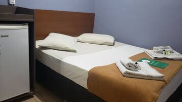 Quarto de hotel com uma cama, uma pequena mesa com frigorífico e uma janela.