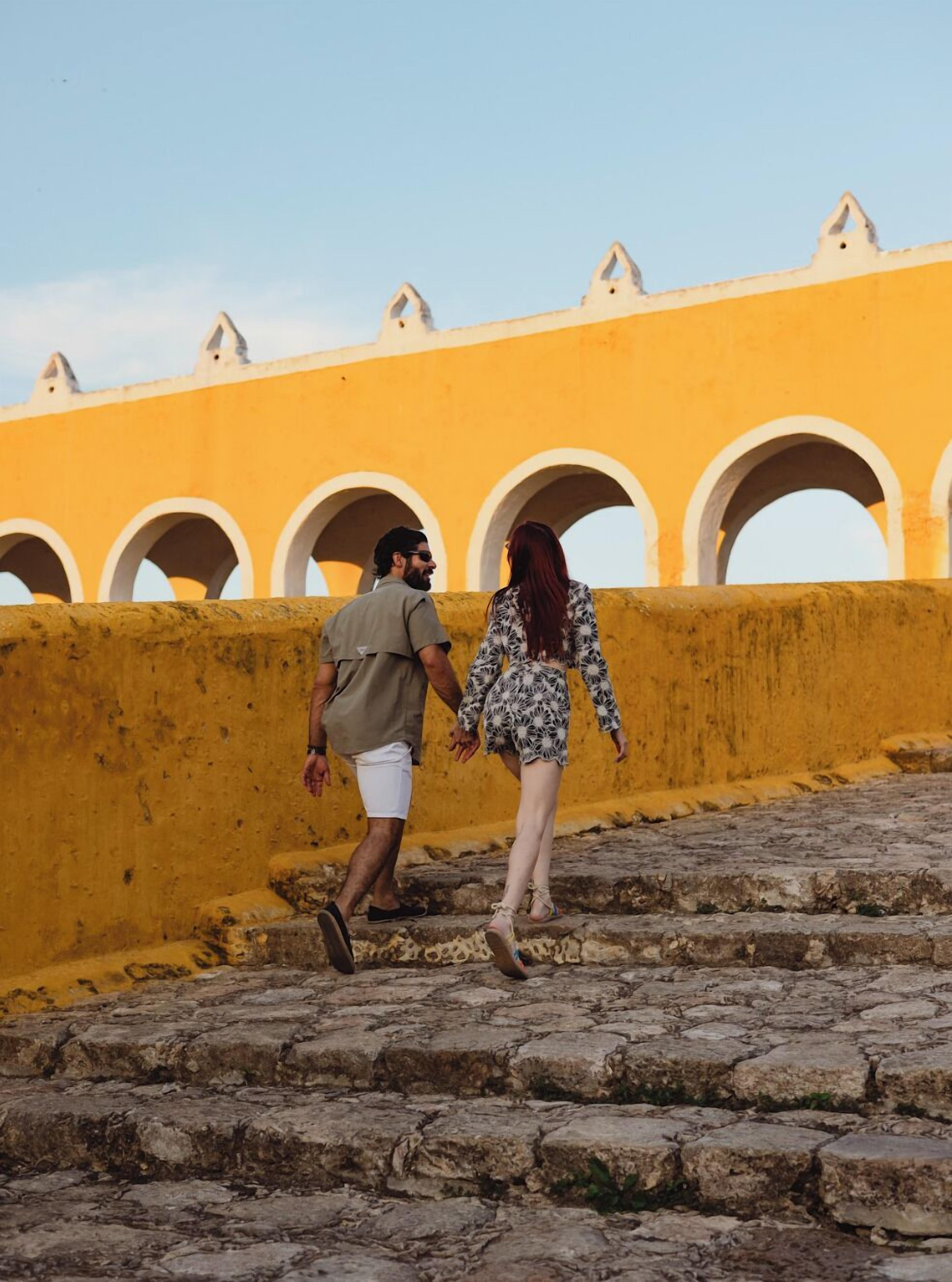 Um casal caminhando em degraus de pedra com uma parede amarela e arcos ao fundo.