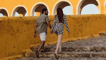 Um casal caminhando em degraus de pedra com uma parede amarela e arcos ao fundo.